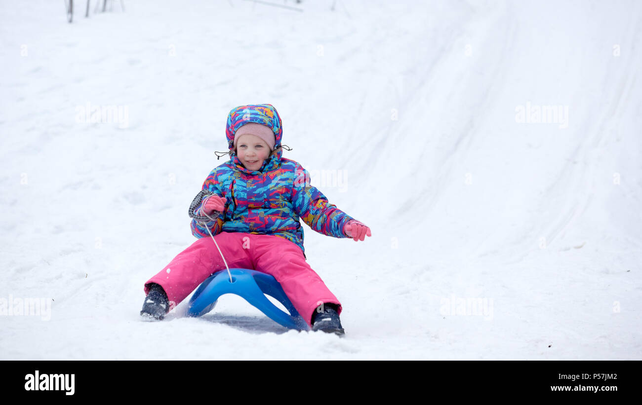 Cheerful girl riding a sled downhill on a snow covered sledge trail in ...