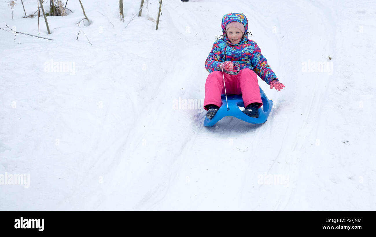 Cheerful girl riding a sled downhill on a snow covered sledge trail in ...