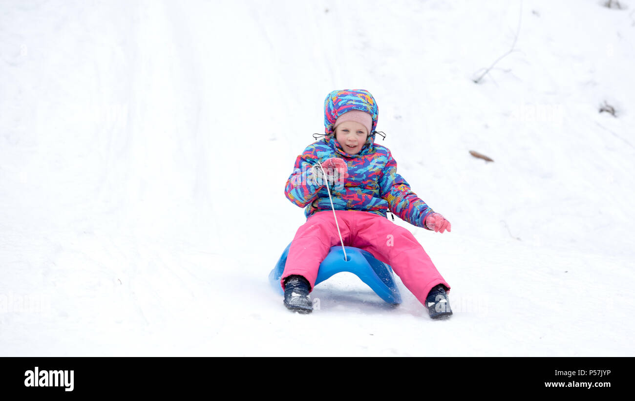 Cheerful girl riding a sled downhill on a snow covered sledge trail in ...