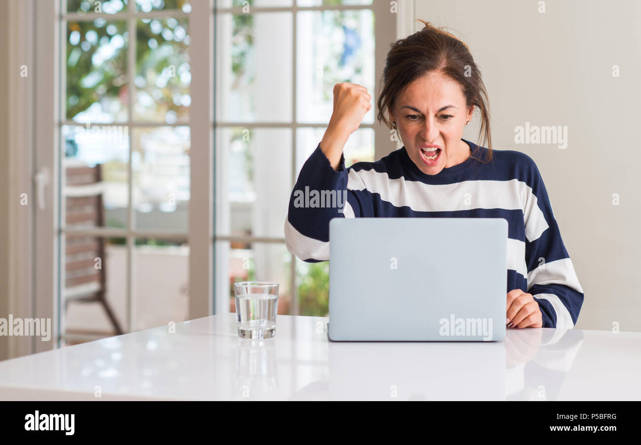 Middle aged woman using laptop at home annoyed and frustrated shouting ...