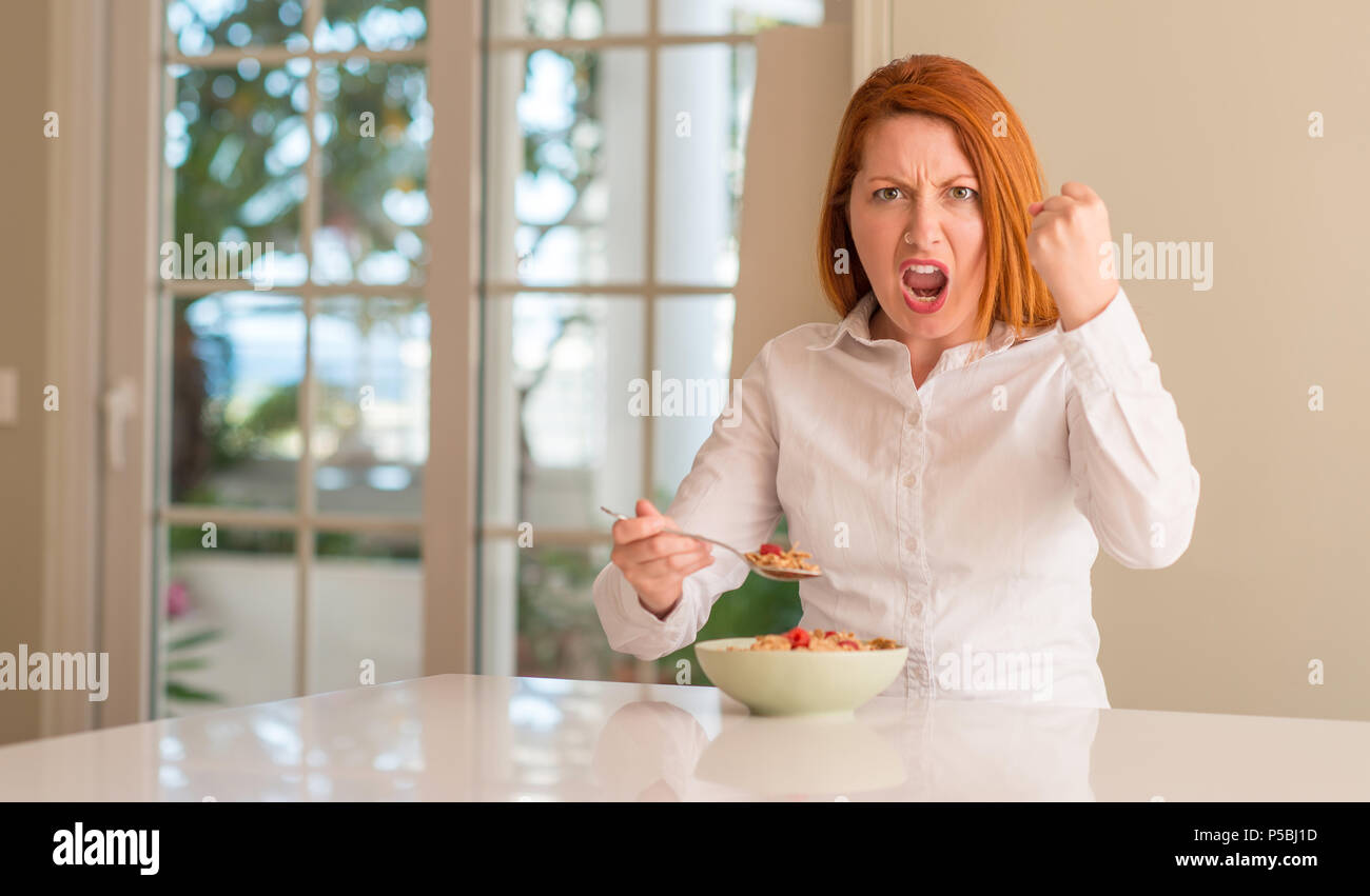 Redhead woman eating cereals with raspberries at home annoyed and ...