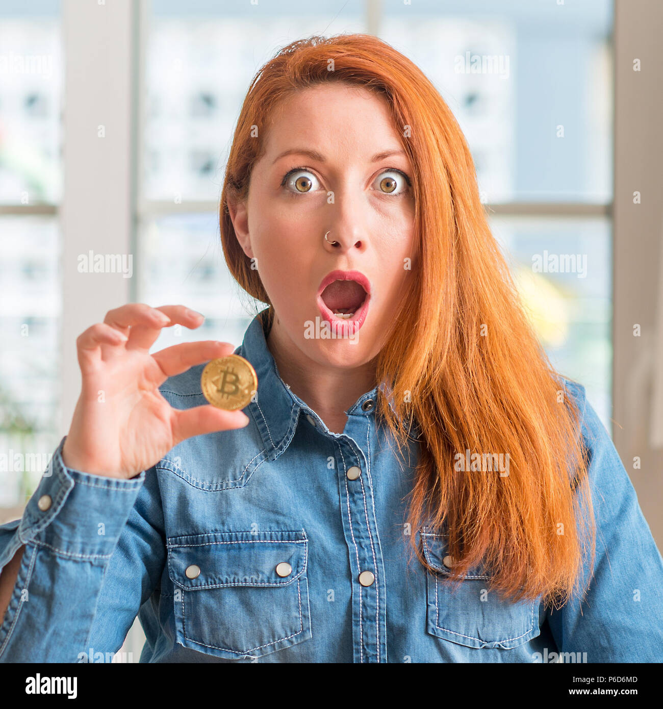 Redhead woman holding bitcoin cryptocurrency at home scared in shock ...