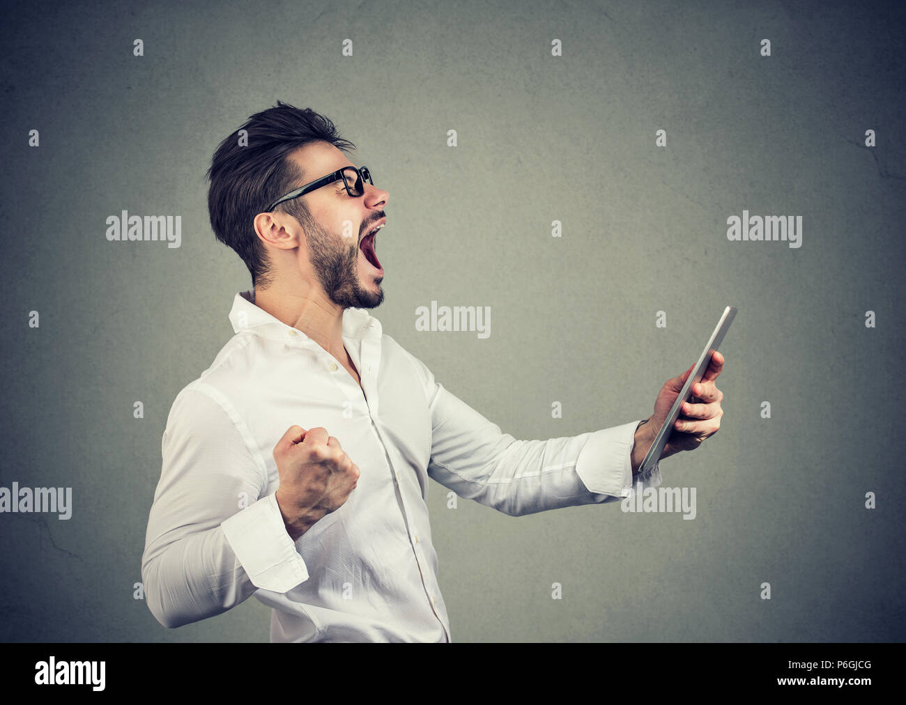 Cheerful hipster man holding tablet and fist up while screaming with ...