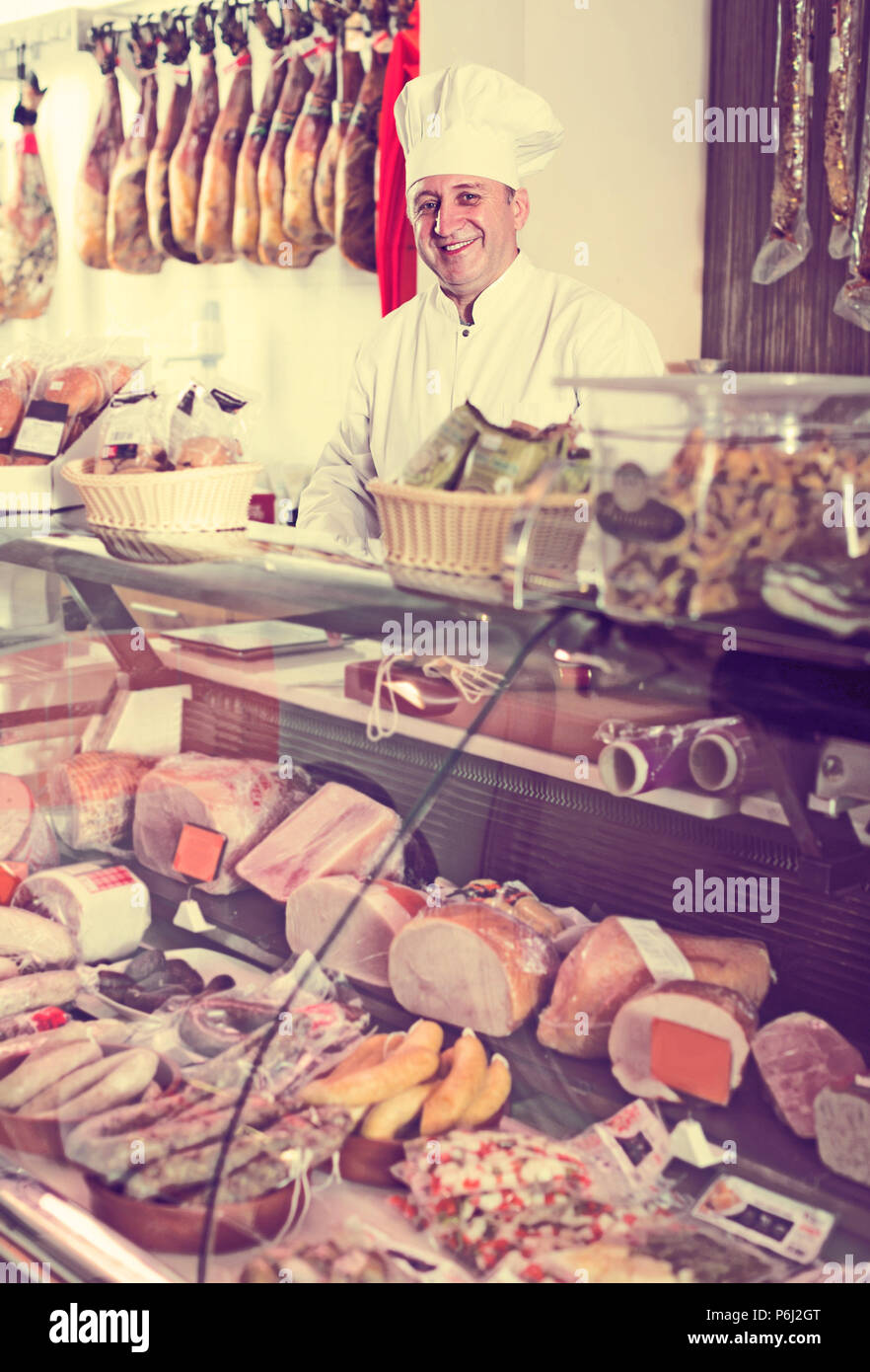 Mature male butcher with wurst, dry-cured and smoked meat at counter ...
