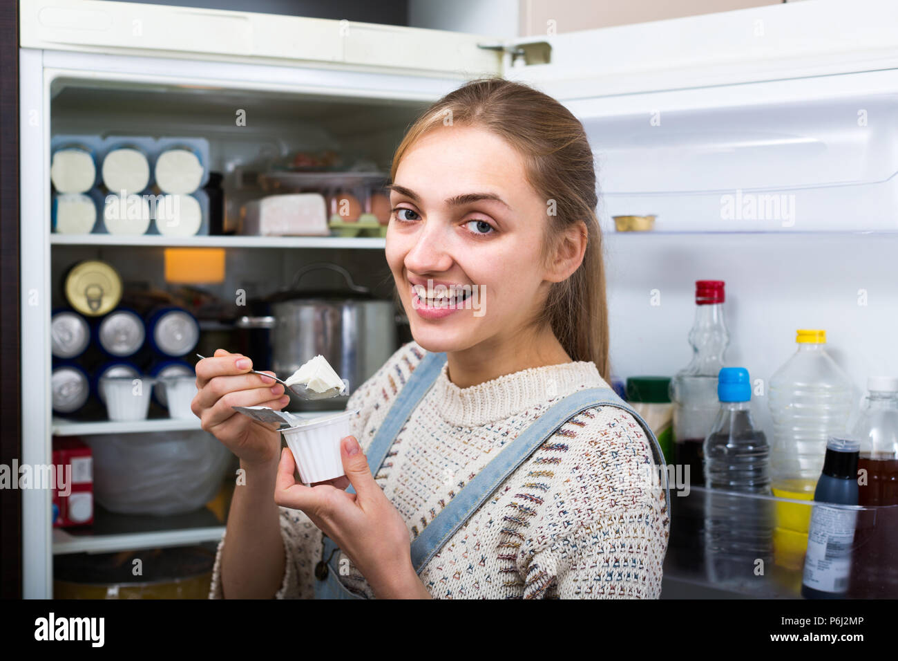 Cheerful young housewife eating sour cream near opened refrigerator ...