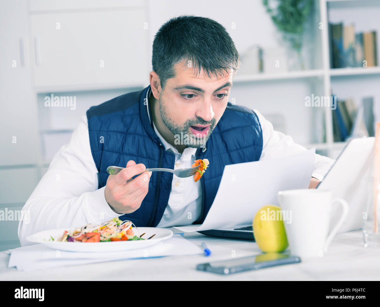 Young man eating vegetable salad and working at laptop at table indoors ...