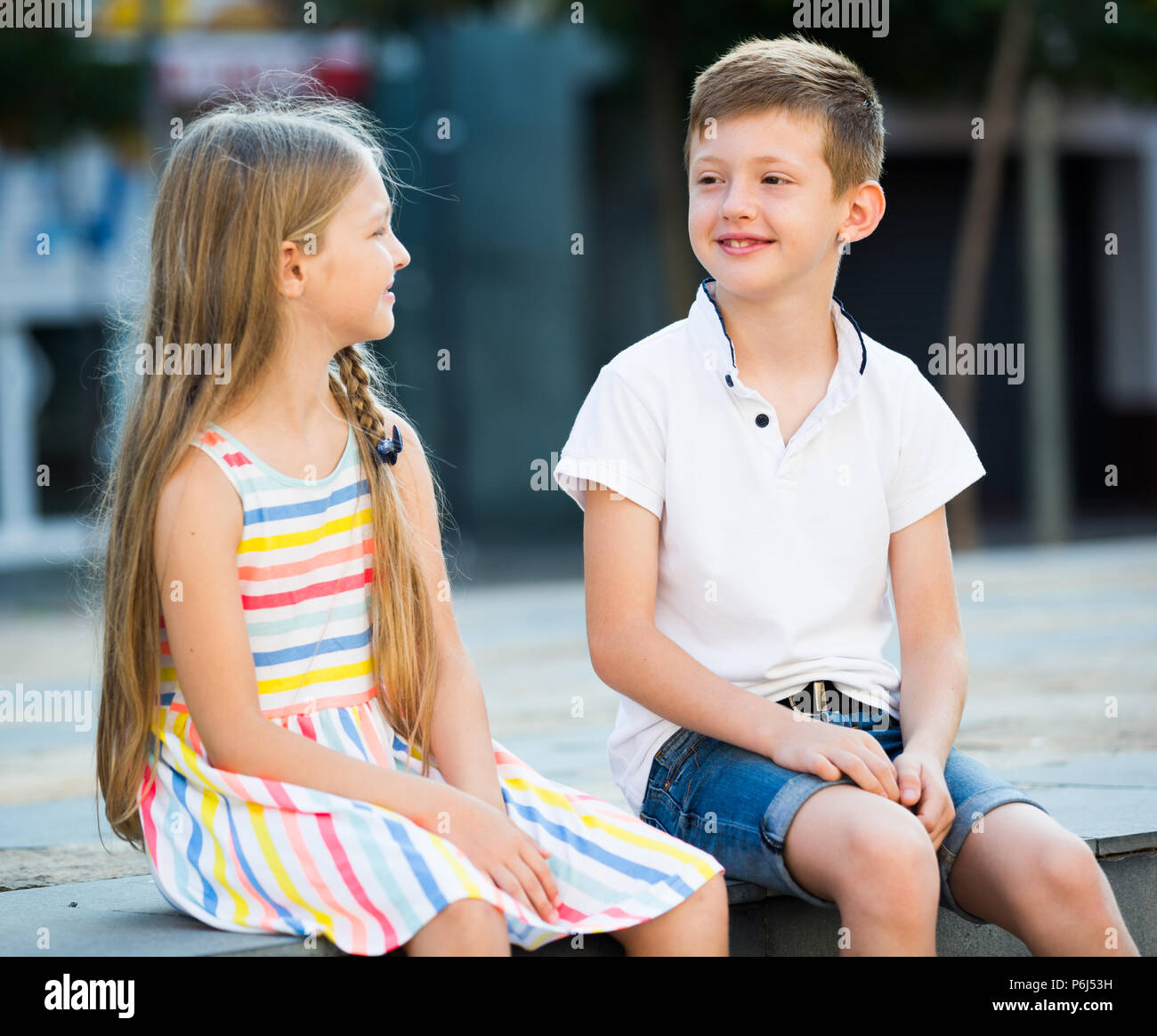 Two smiling kids in preschool age sitting together outdoors in town ...