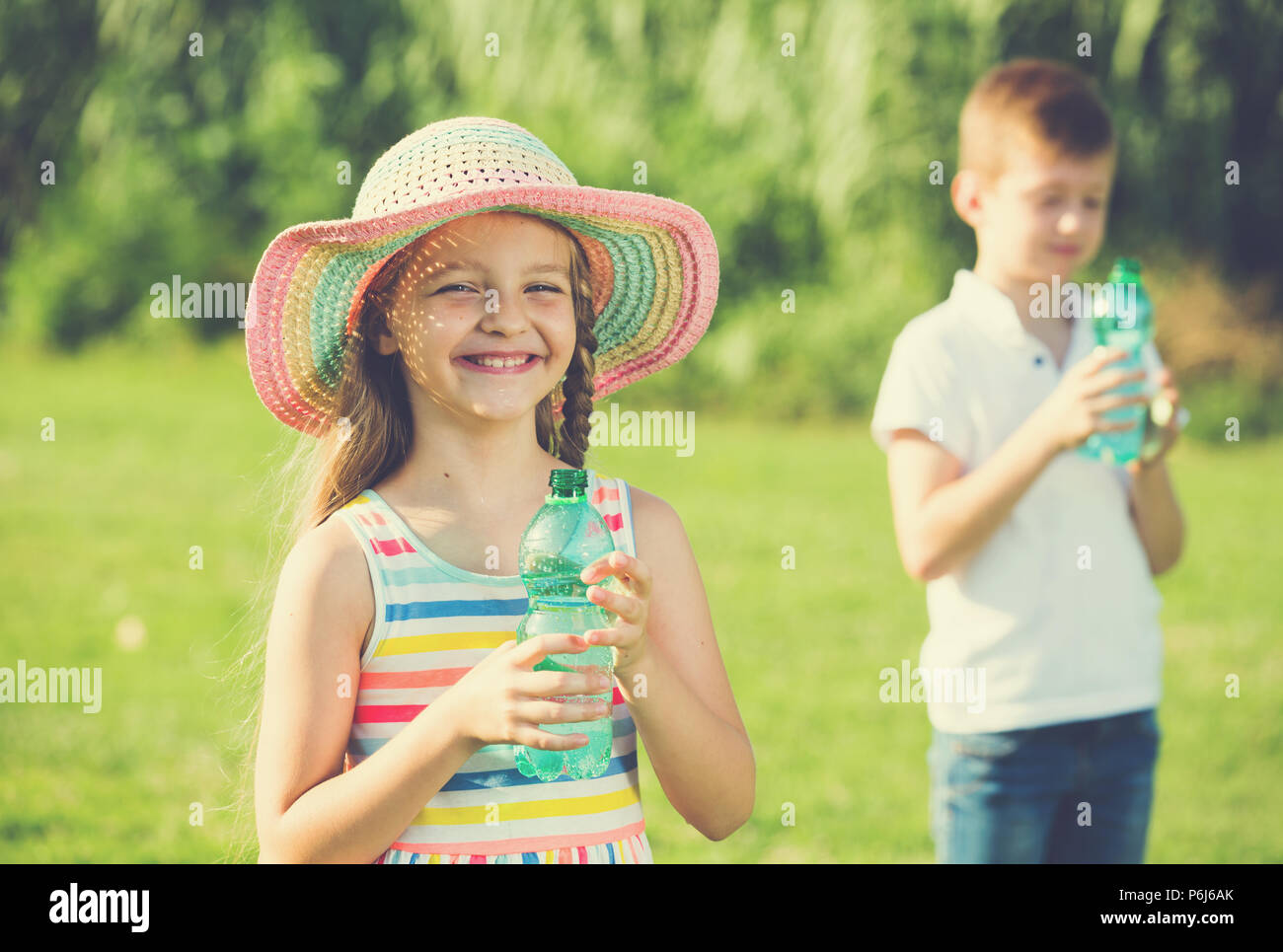 smiling girl in bright dress and sun hat drinking water on warm summer ...