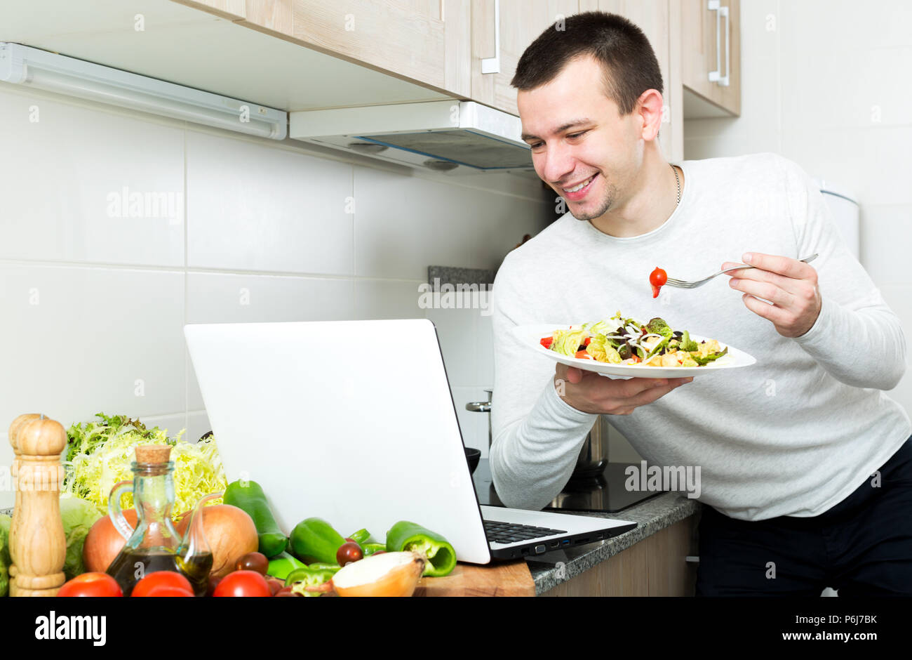 Handsome adult man tasting vegetables dish near laptop in home interior ...