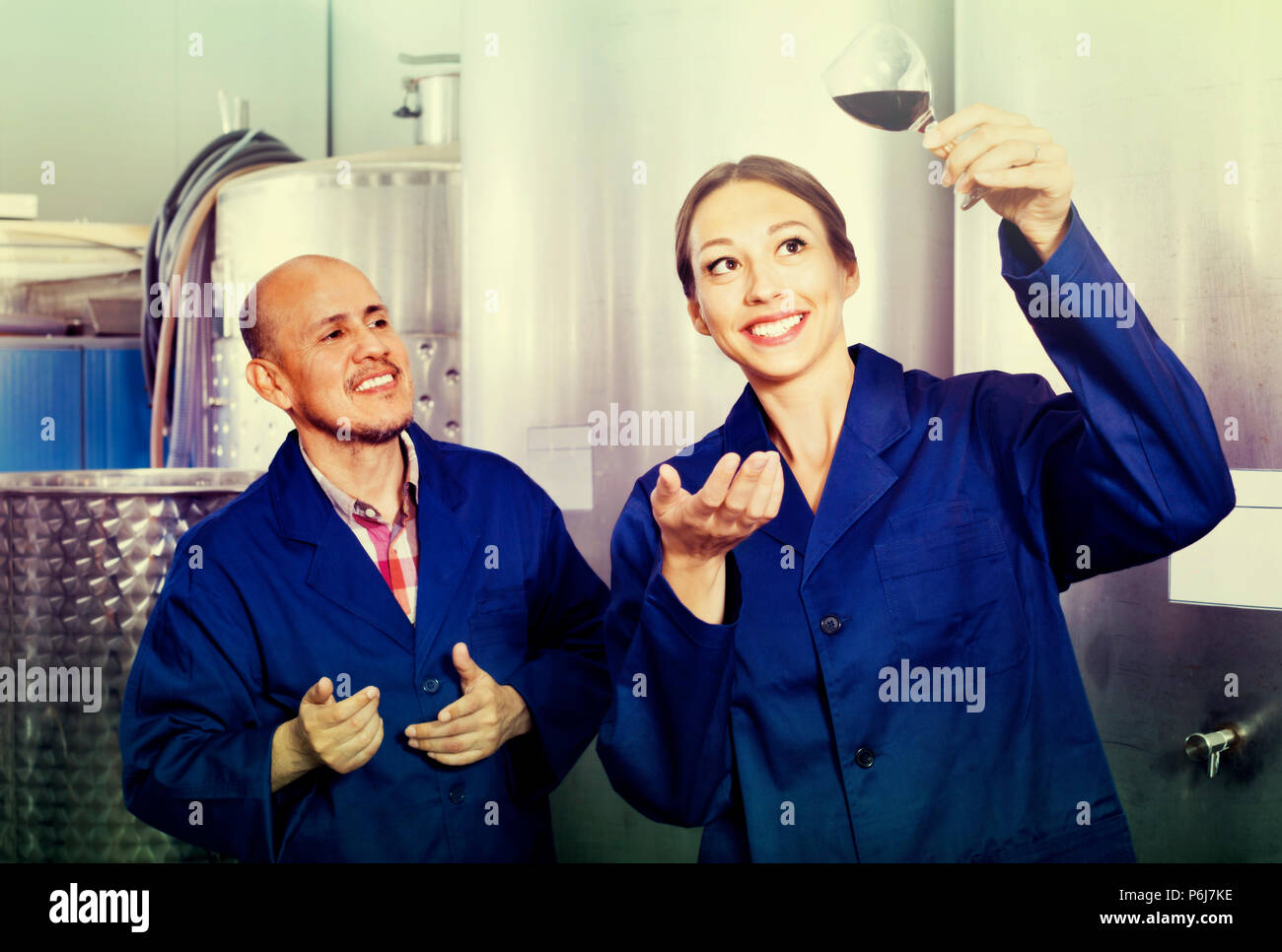 Two cheerful winery workers holding glass of wine in fermenting section ...