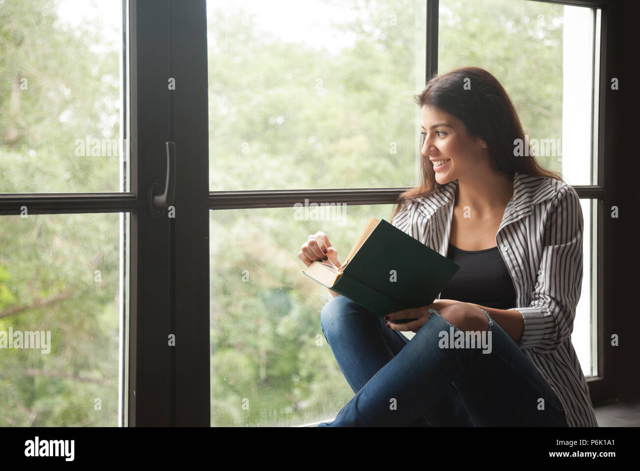 Smiling girl distracted from reading novel looking in window Stock ...