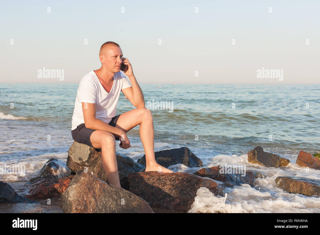 Young man sitting on a rock by the sea and talking on the phone Stock ...