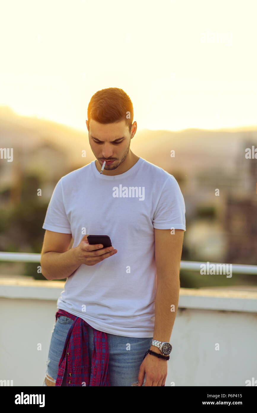 Young man holding his cellphone while smoke cigarette on the balcony in ...