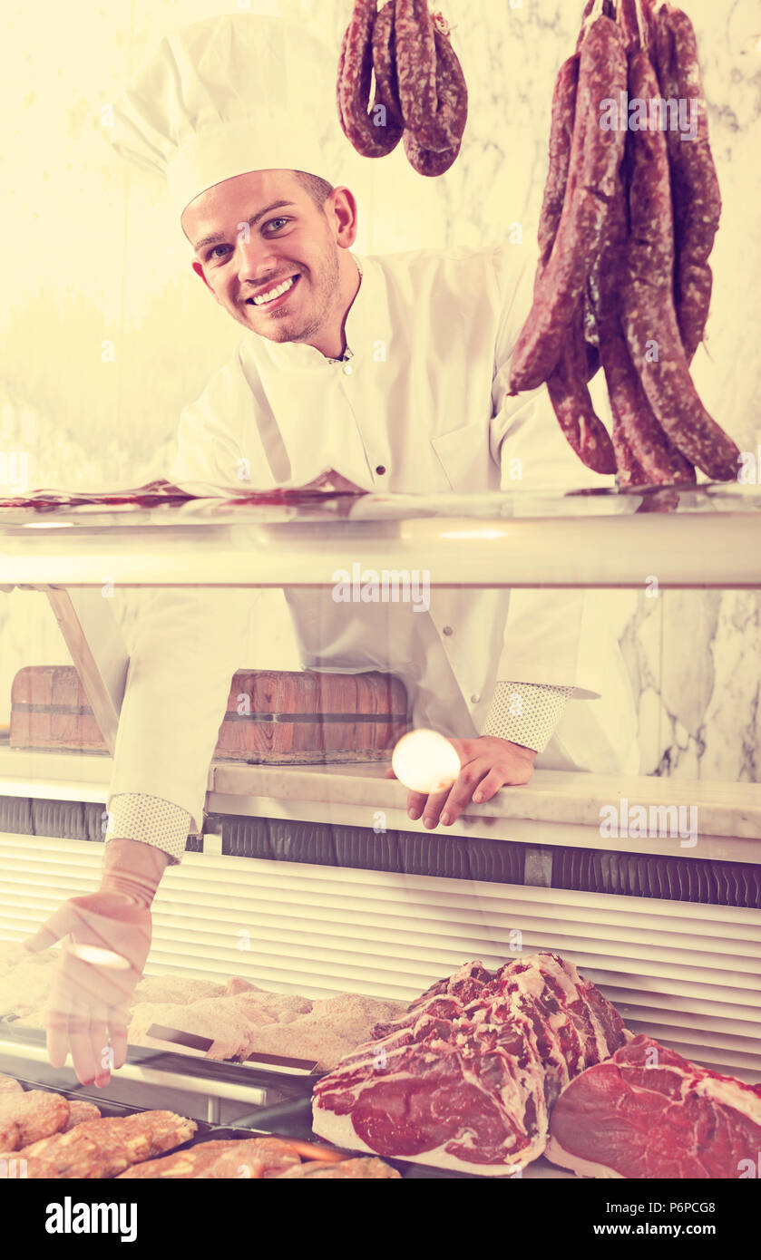 Smiling young cook in white coat at butcher store offering meat and ...
