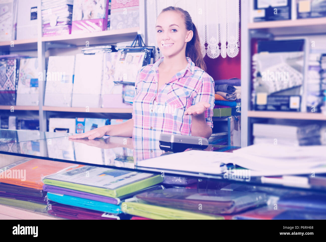 Positive female shop assistant offering help at counter of textile shop ...
