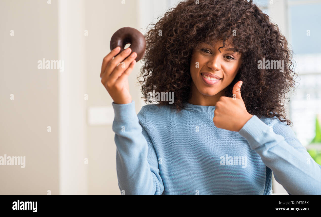 Young african american woman holding chocolate donut at home happy with ...