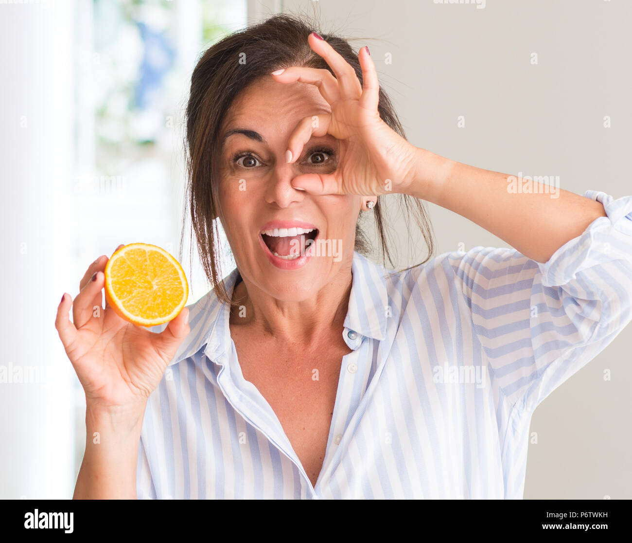 Middle aged woman holding orange fruit with happy face smiling doing ok ...