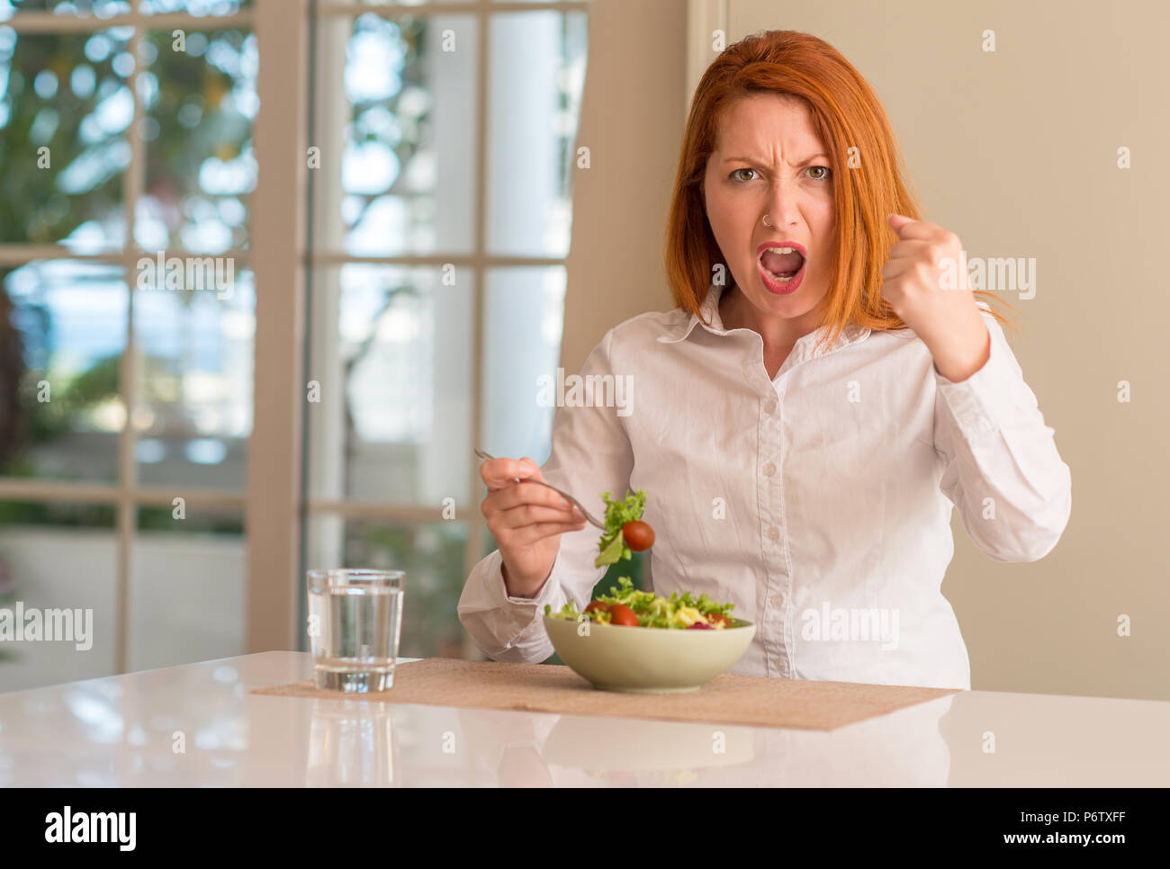 Redhead woman eating fresh green salad at home annoyed and frustrated ...