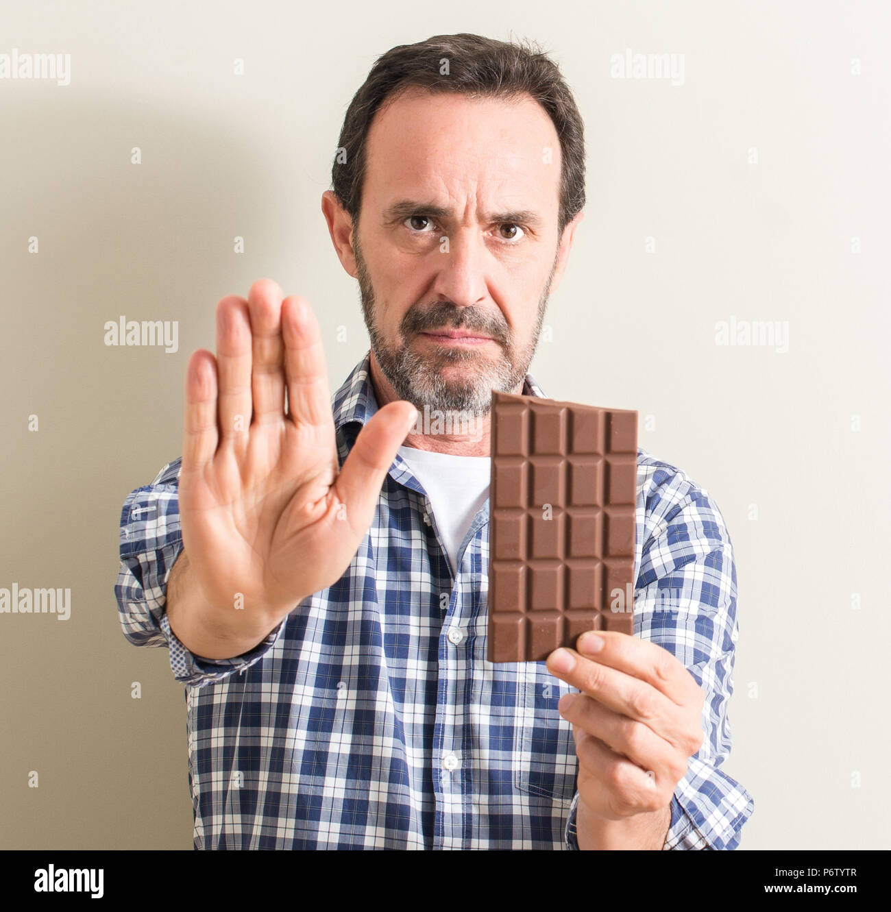 Senior man eating chocolate bar with open hand doing stop sign with ...