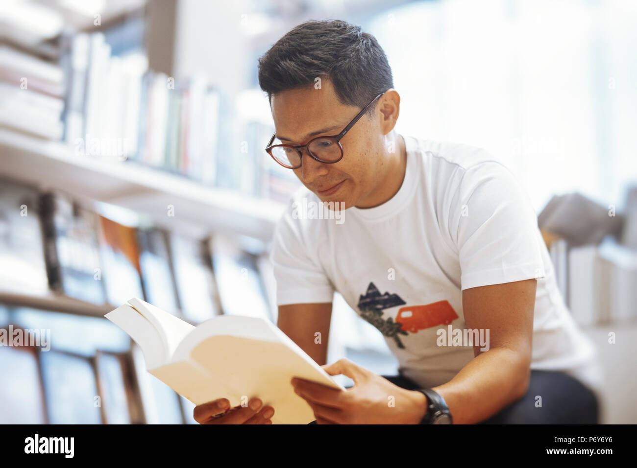 Young spectacled man sitting in a book store and reading book with ...