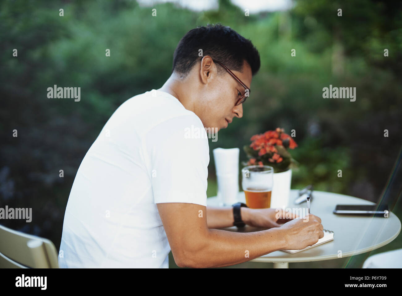 Young spectacled man drinking cold beer and writing some notes in ...