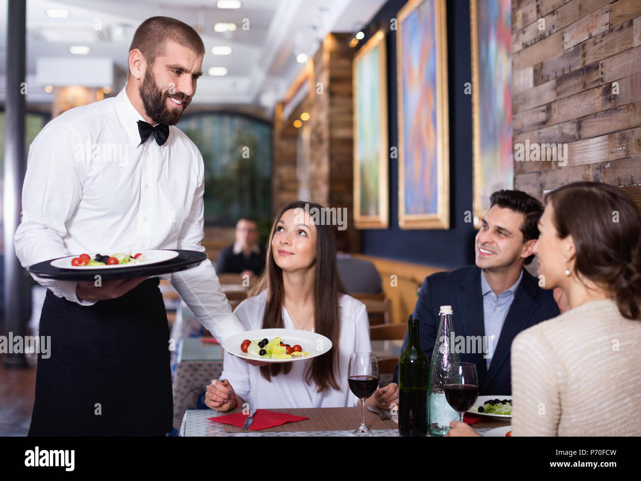 Diligent friendly smiling waiter with dishes serving man and woman ...