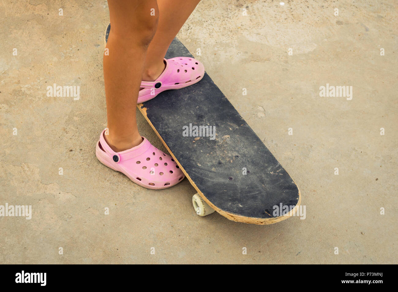 Little girl wearing pink plastic clogs riding on a skateboard. Learning ...