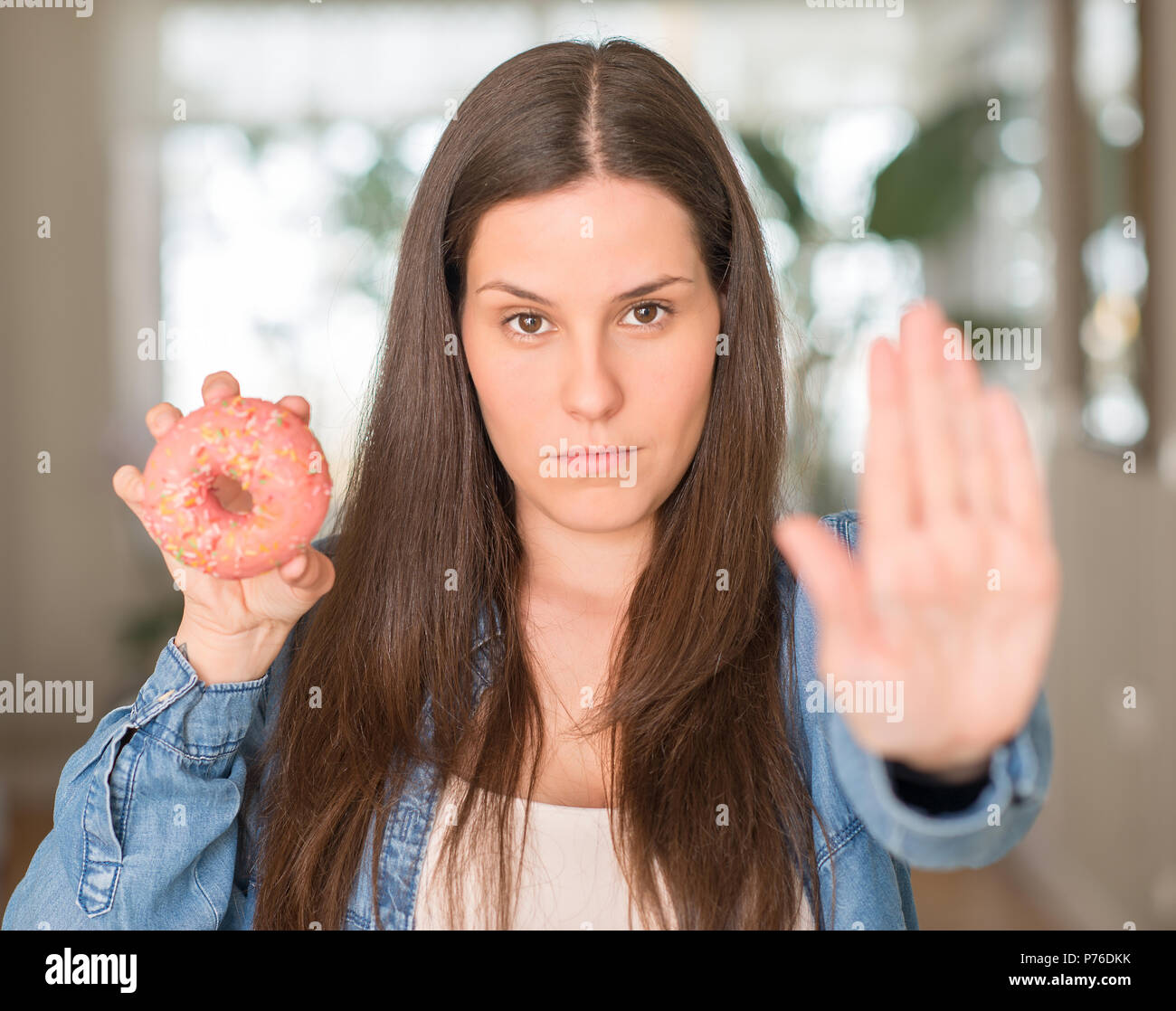 Hungry young woman holding pink donut with open hand doing stop sign ...