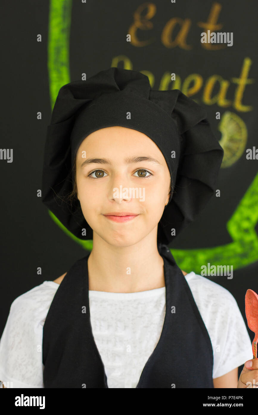 Beautiful young girl cook chef in a black chef's cap looks directly to ...