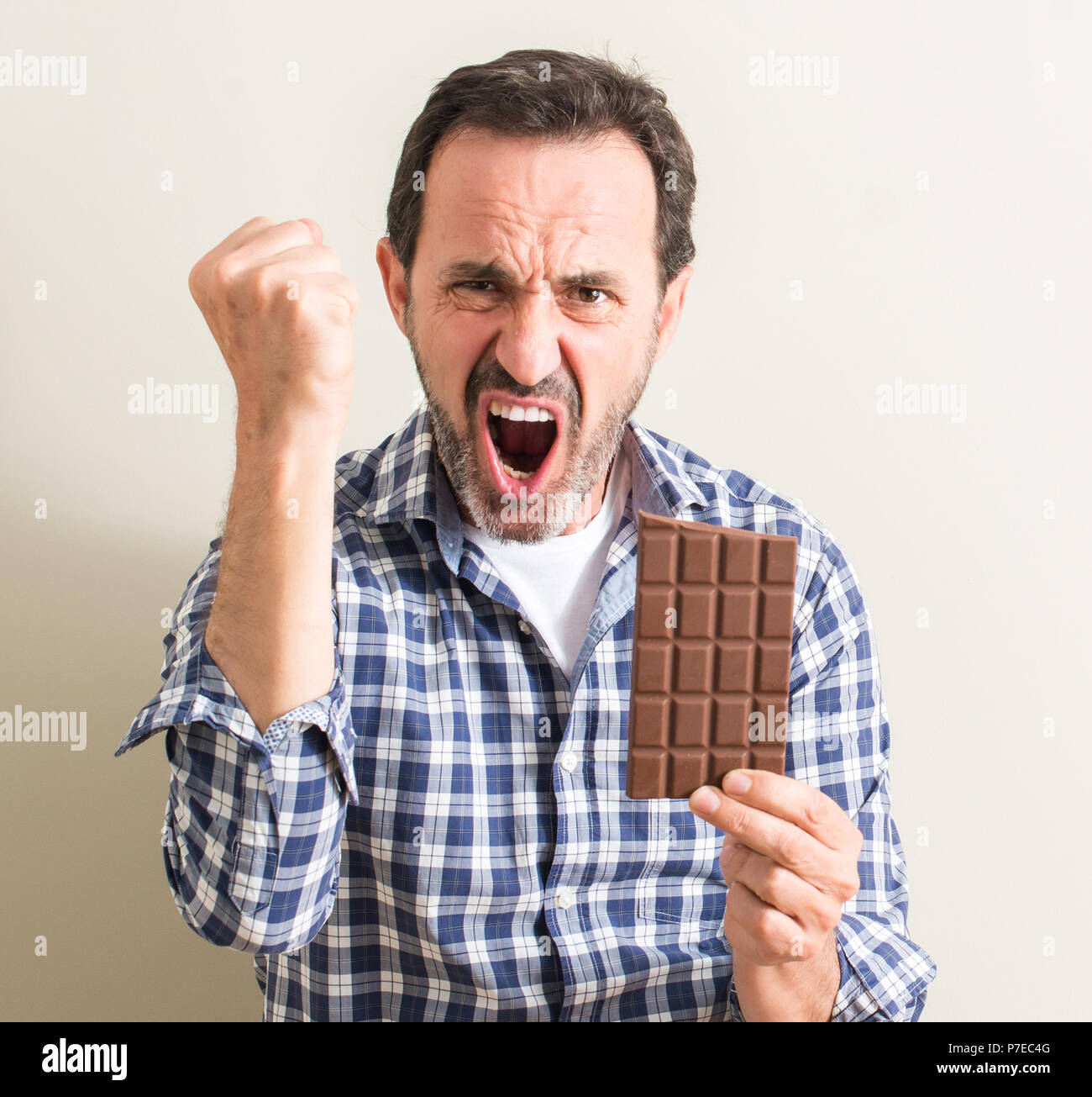 Senior man eating chocolate bar annoyed and frustrated shouting with ...