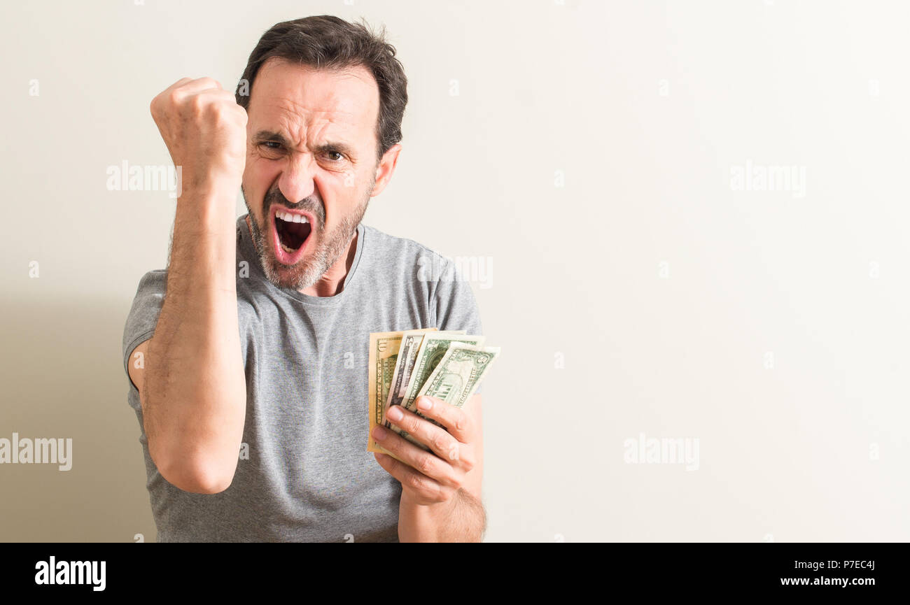 Senior man holding dollar money annoyed and frustrated shouting with ...