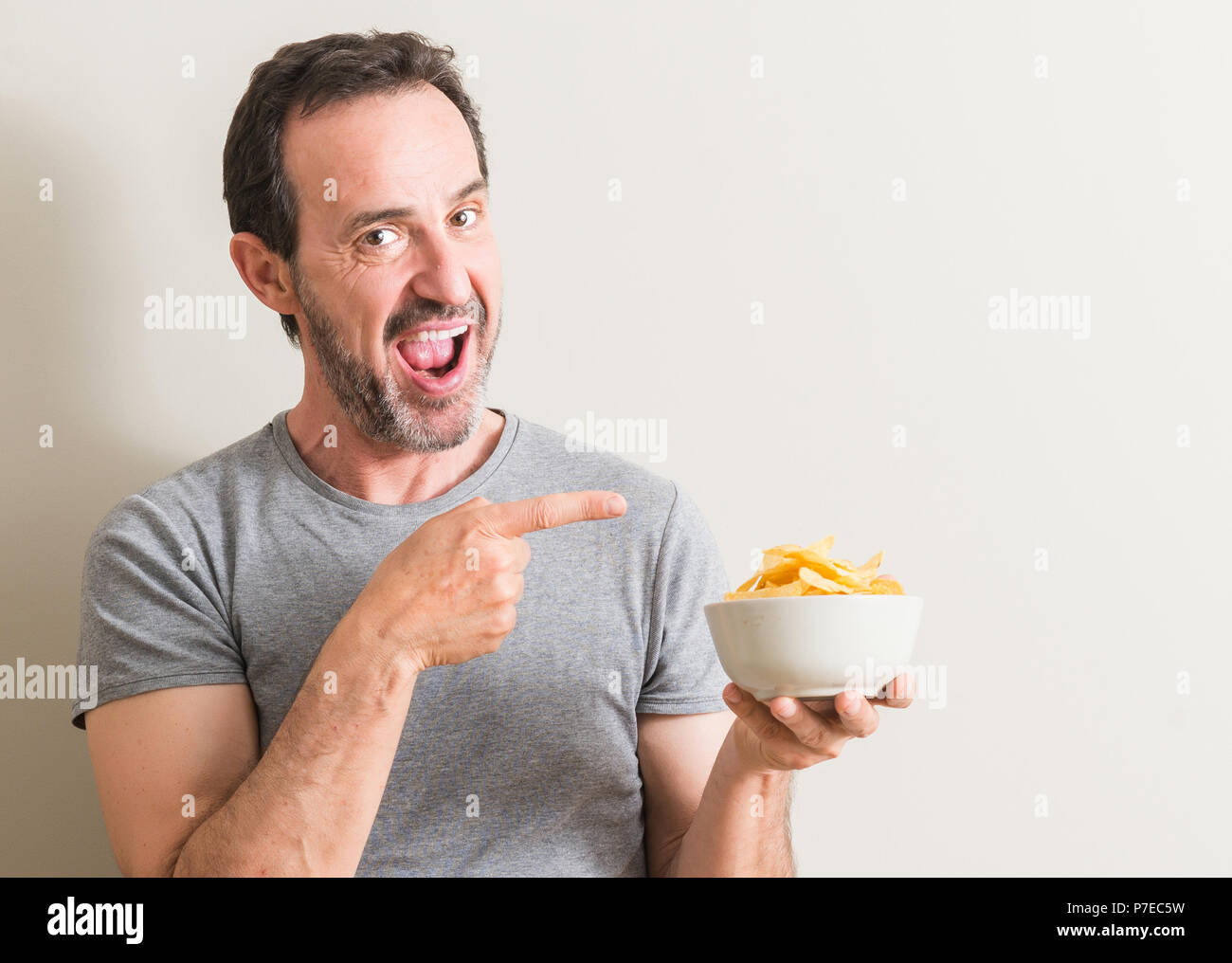 Senior man eating potato chips very happy pointing with hand and finger ...
