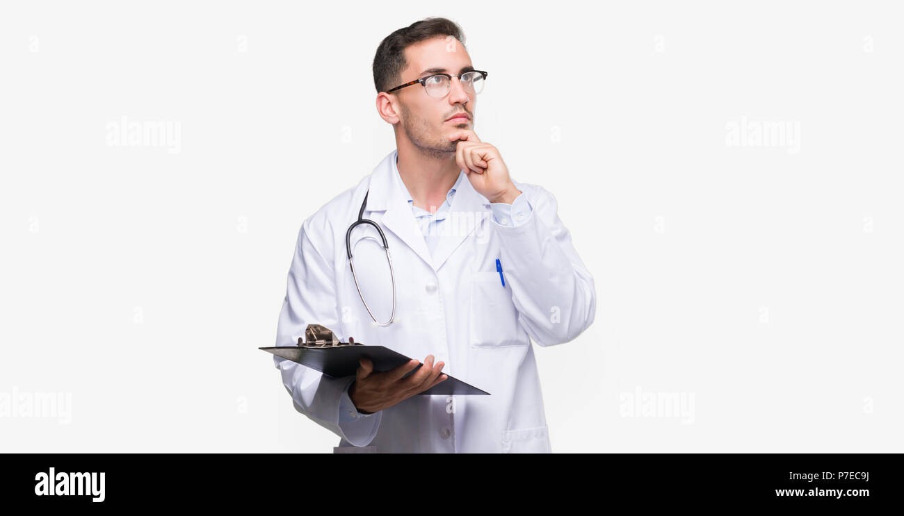 Handsome young doctor man holding a clipboard serious face thinking ...