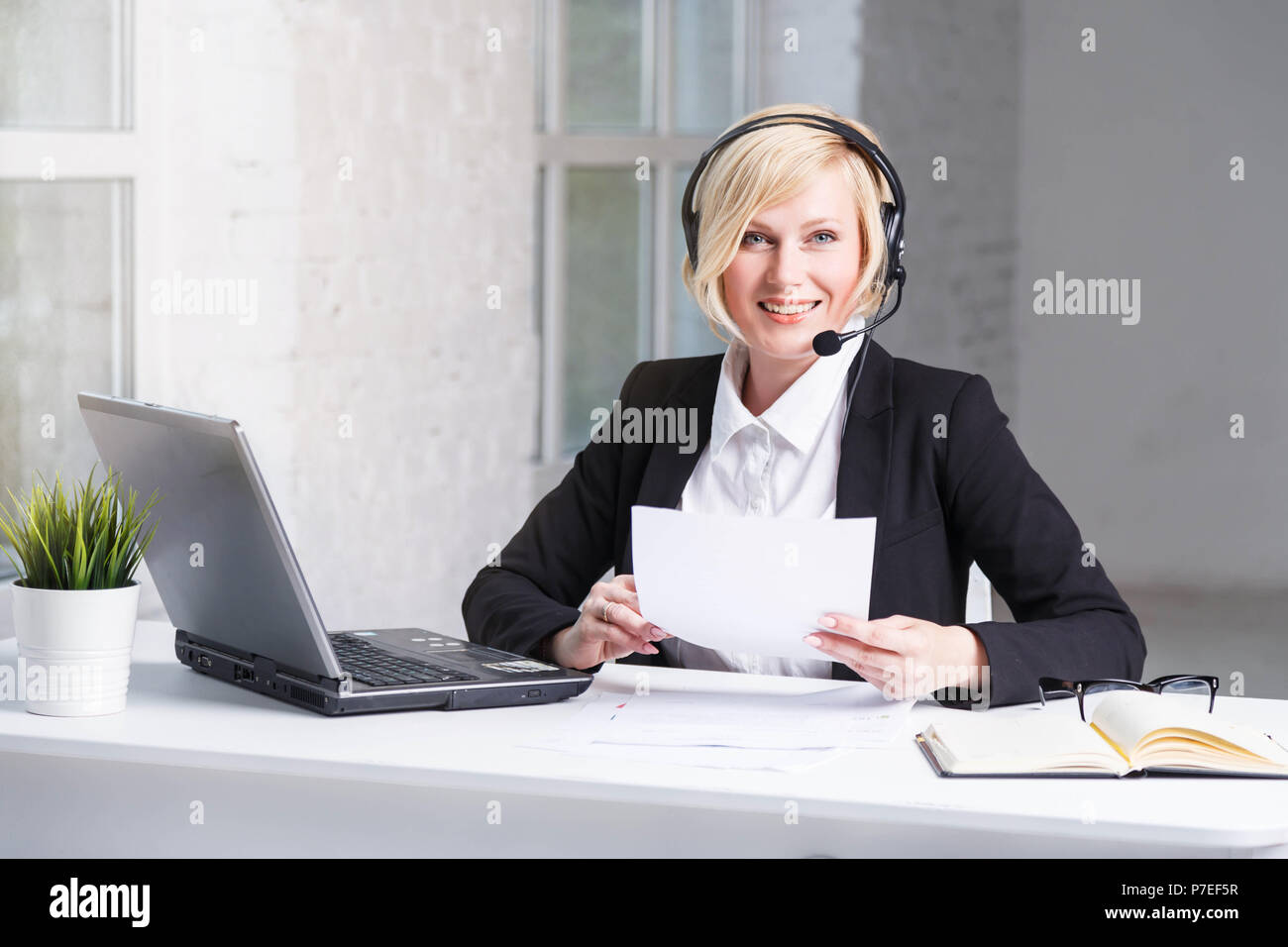 Beautiful smiling blonde woman, call center operator dressed in black stylish suit with headset ...