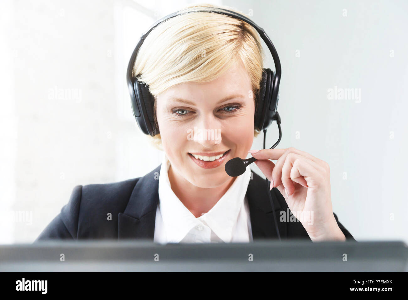 Smiling call center operator, blonde woman dressed in black jacket and white shirt working on ...