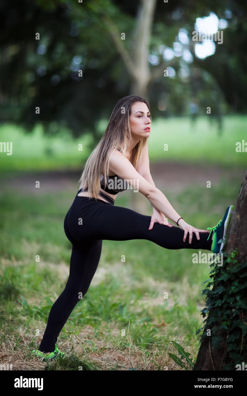 A young woman does stretching exercises leaning on the trunk of a tree ...