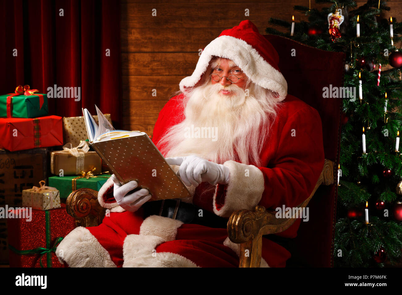 Santa Claus sits in his chair in front of presents and christmas tree ...