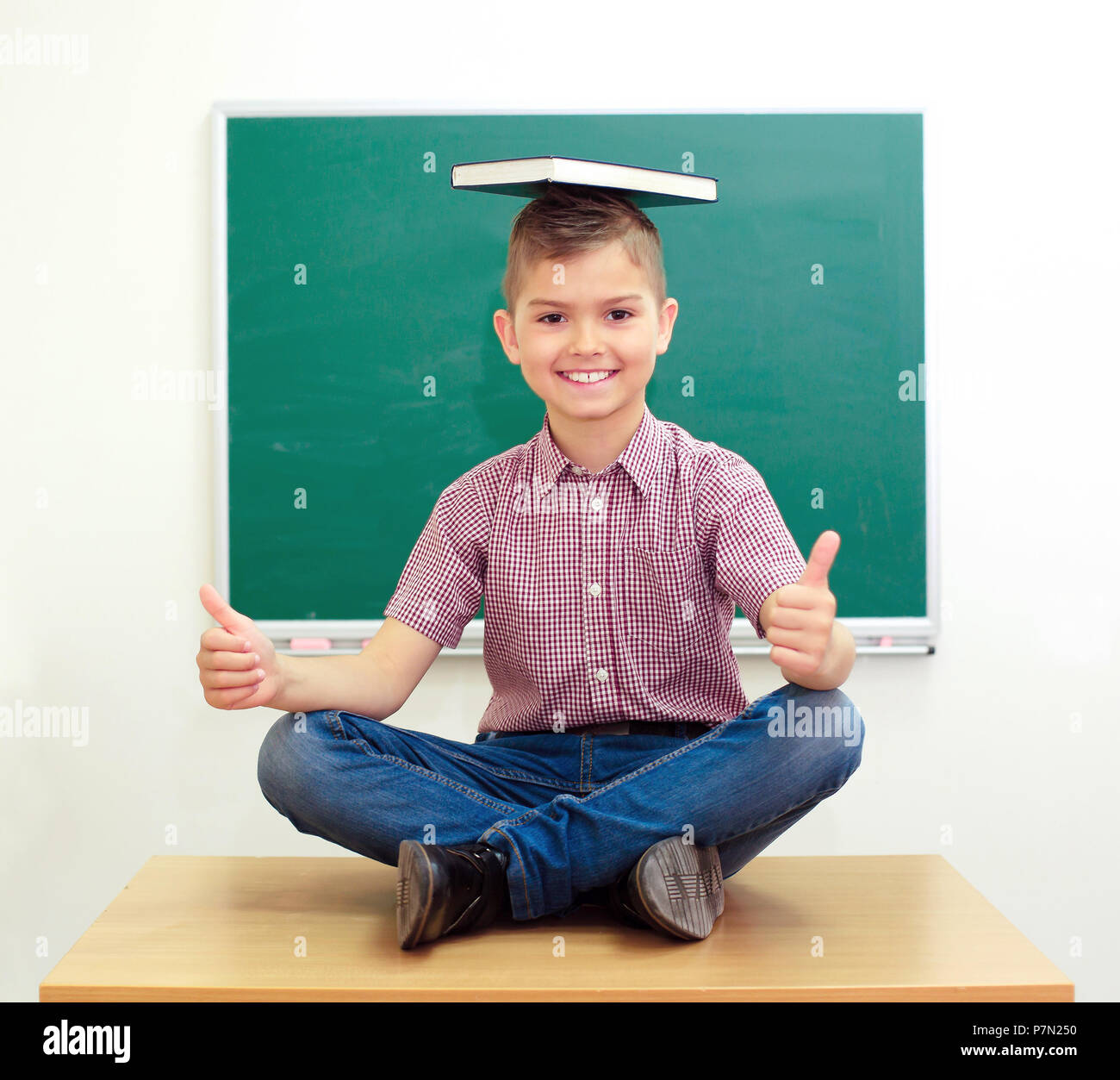 Happy schoolboy with book on head sitting in lotus pose in class room ...