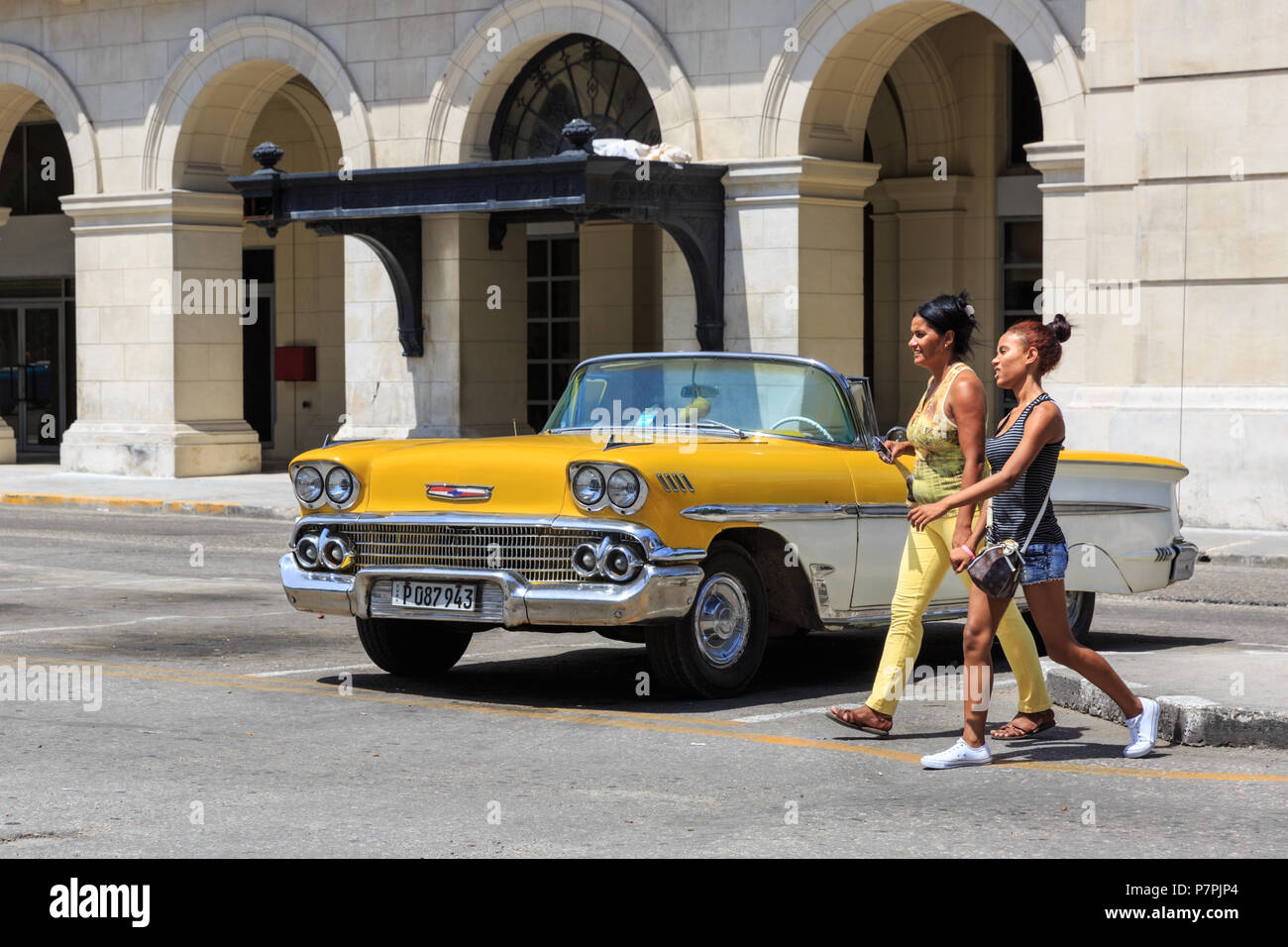 Two Cuban women walk past a striking yellow Chevrolet Impala ...