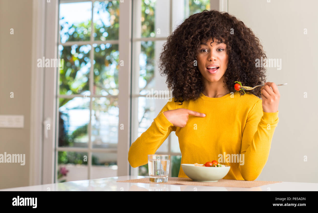 African american woman eating pasta salad with surprise face pointing ...
