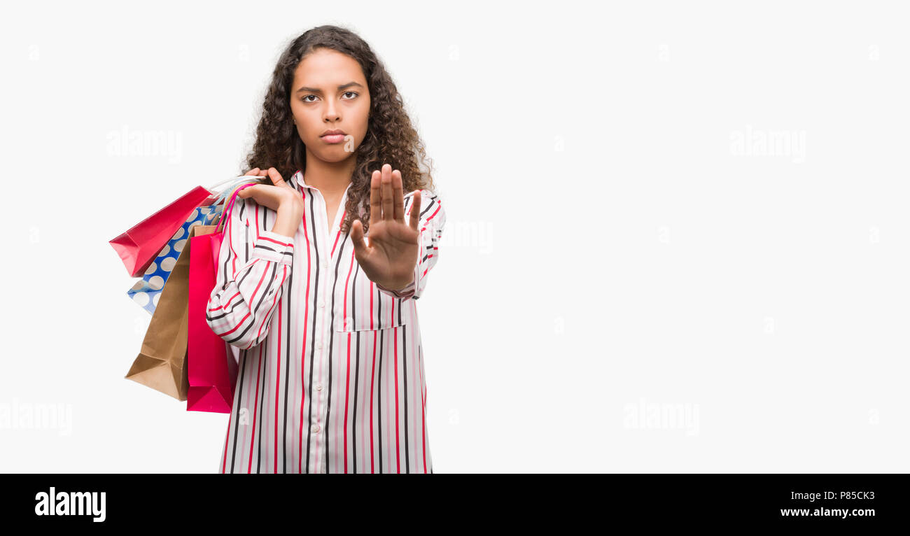 Young hispanic woman holding shopping bags with open hand doing stop ...