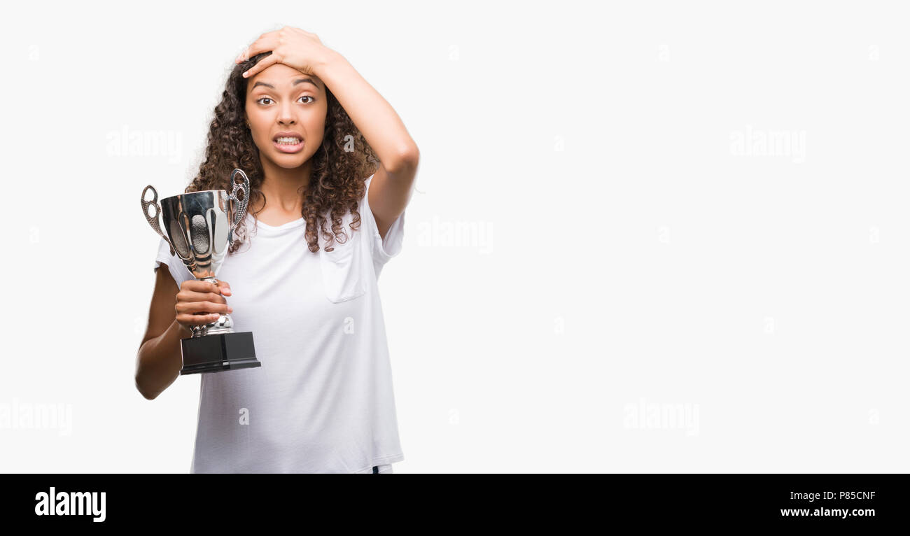 Young hispanic woman holding trophy stressed with hand on head, shocked ...