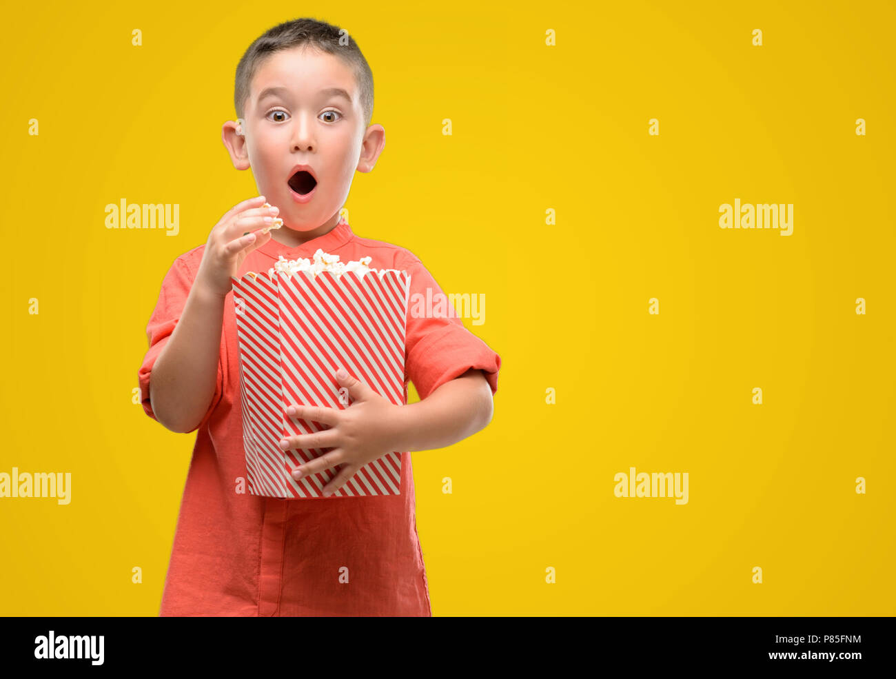 Dark haired little child eating popcorn scared in shock with a surprise ...