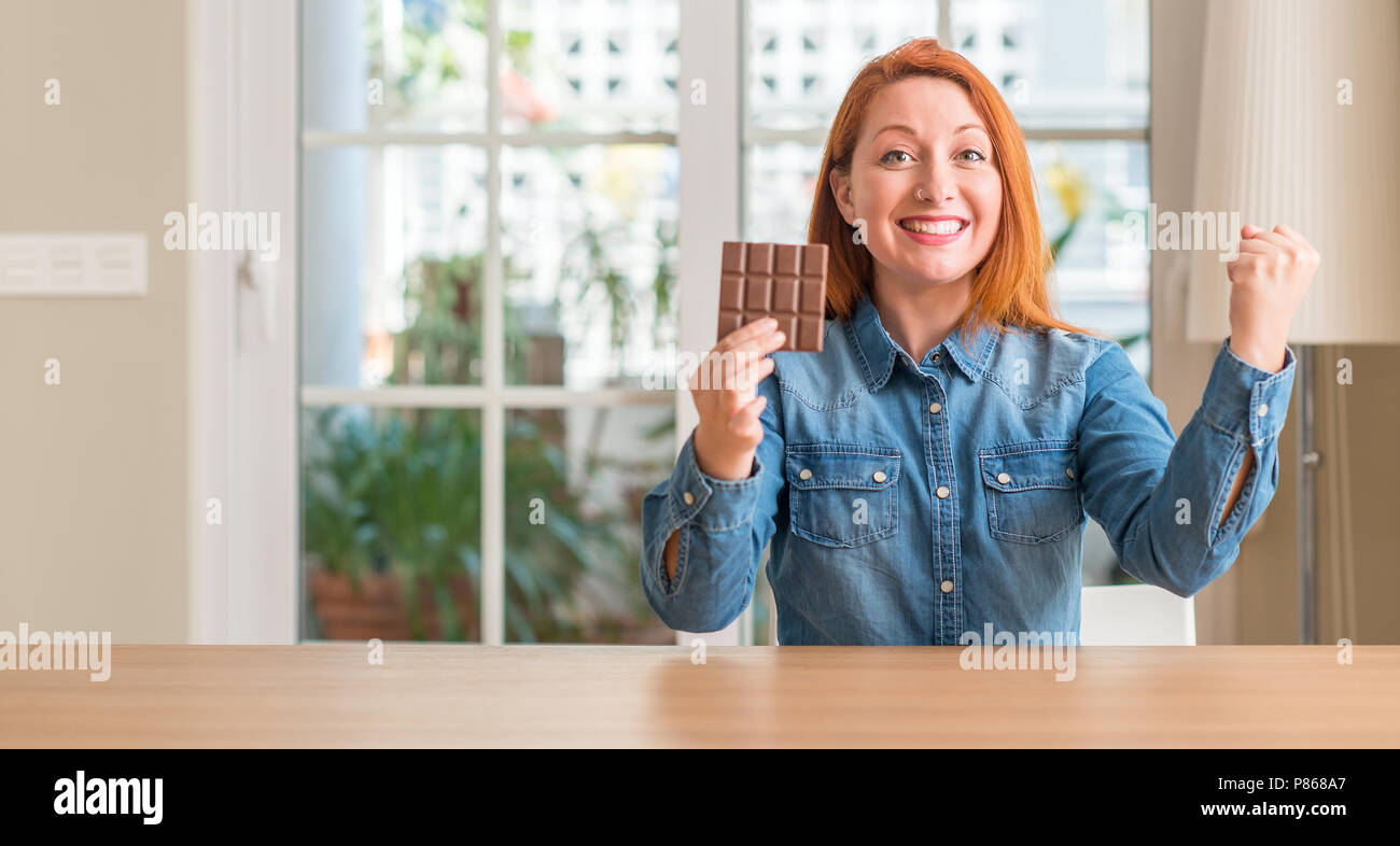 Redhead woman holding chocolate bar at home screaming proud and ...