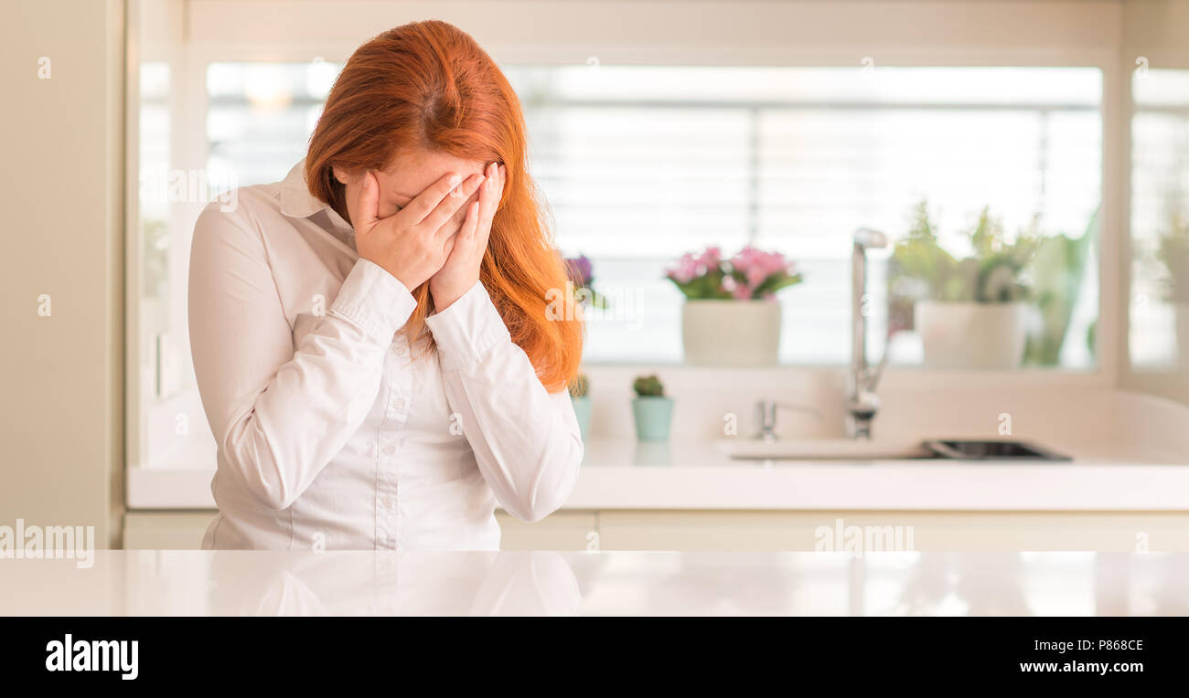 Redhead woman at kitchen with sad expression covering face with hands ...