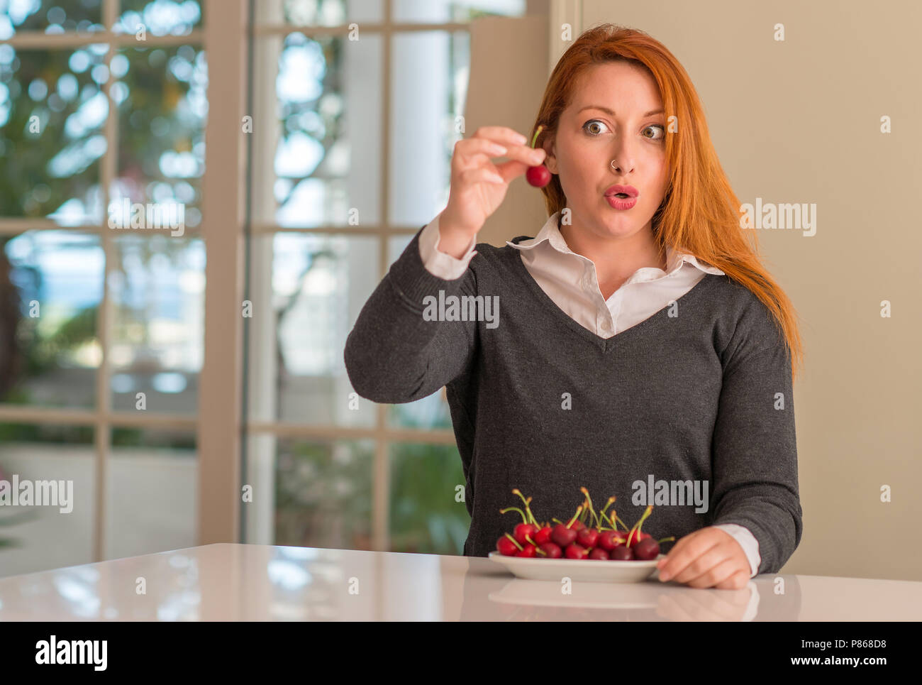Redhead woman eating cherries at home scared in shock with a surprise ...