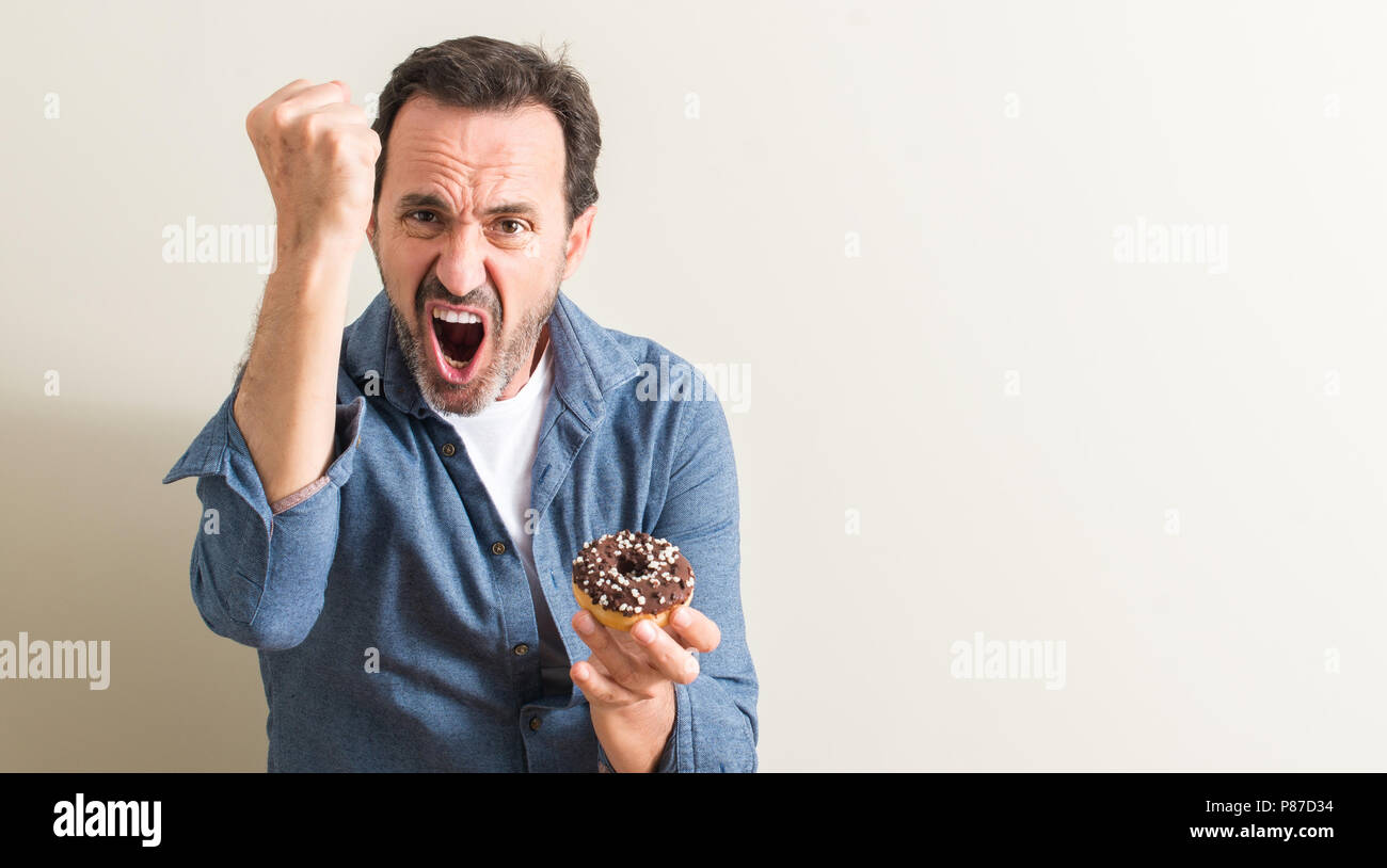 Senior man eating chocolate donut annoyed and frustrated shouting with ...