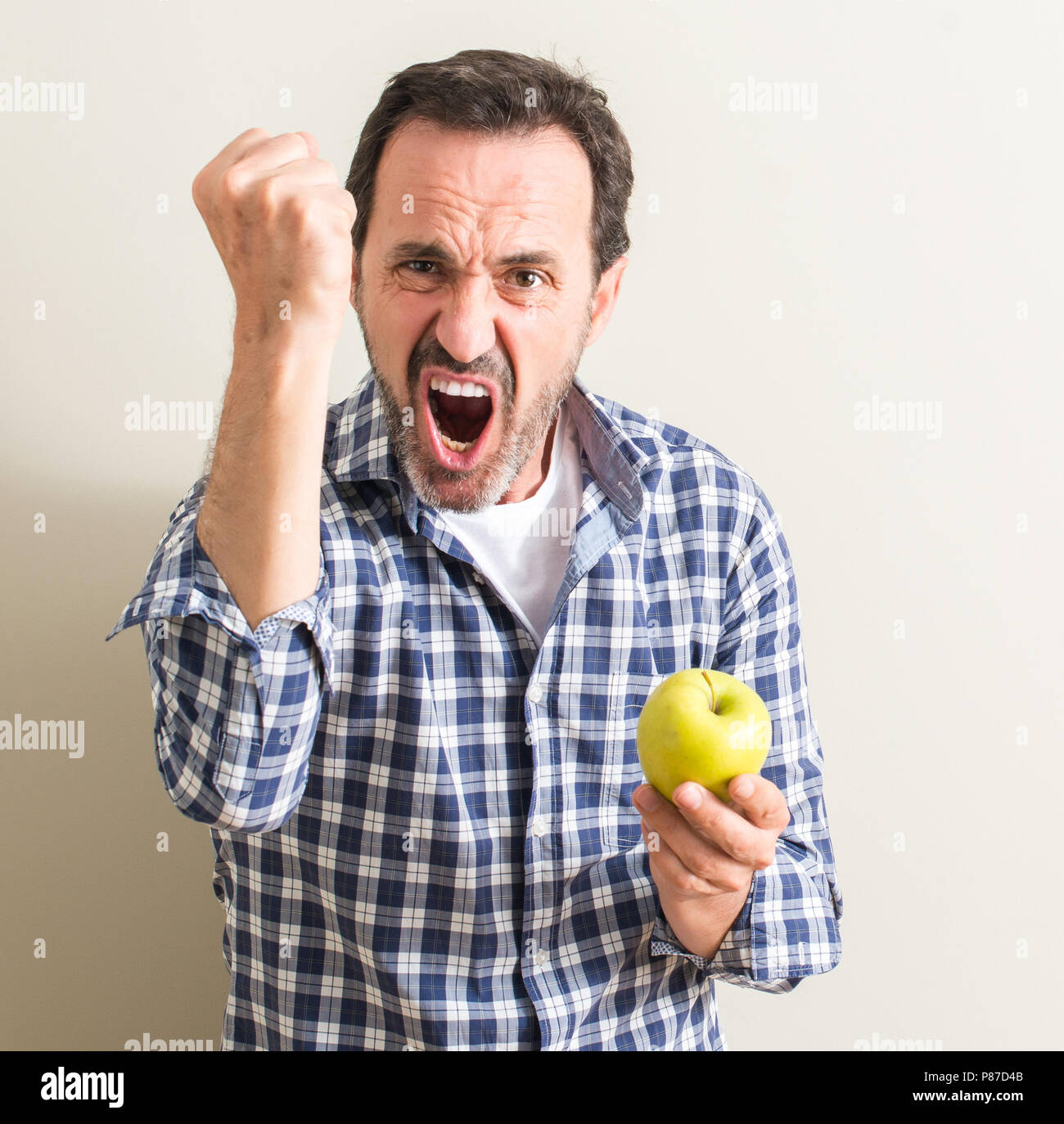 Senior man holding a green apple annoyed and frustrated shouting with ...