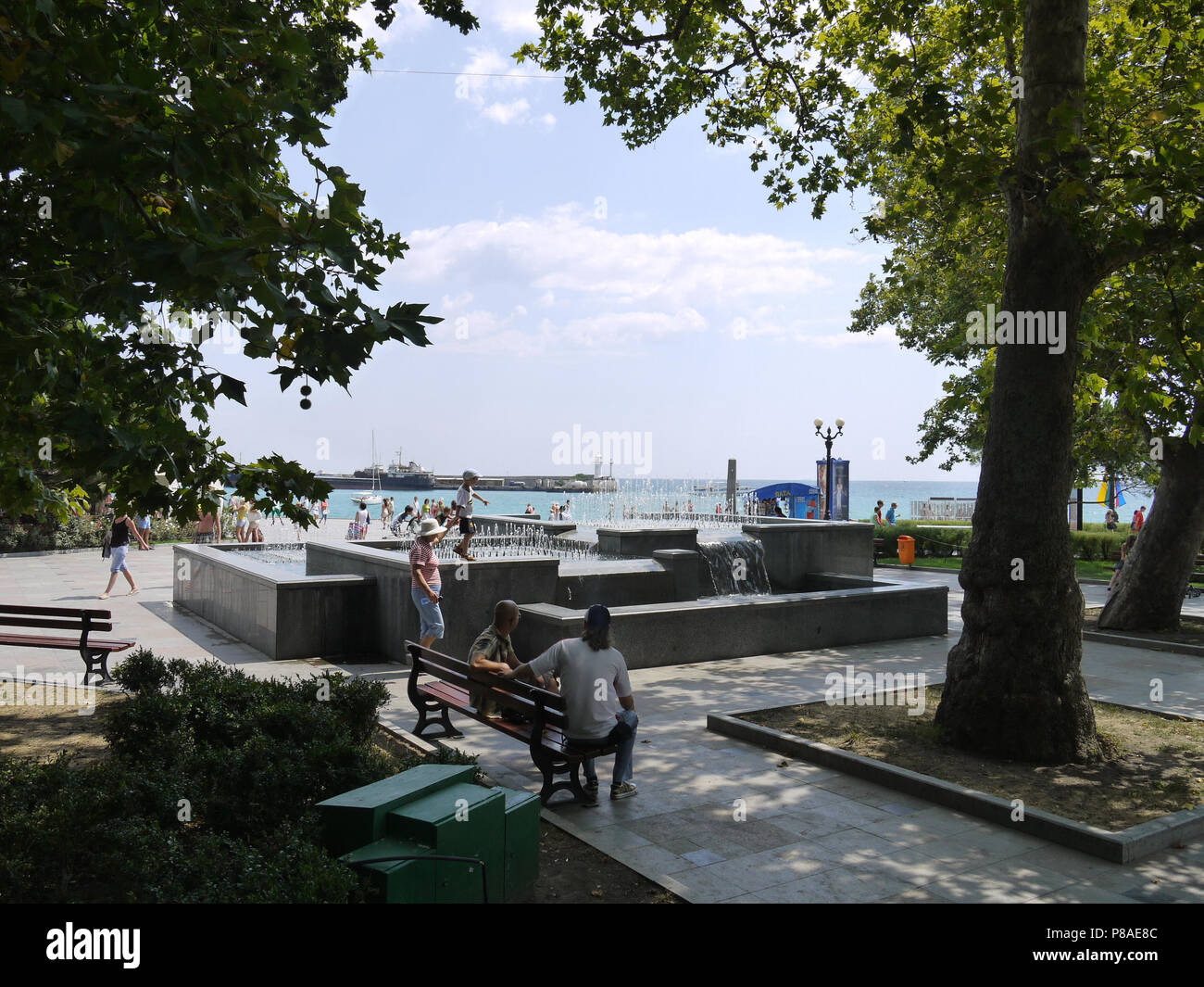people in a park resting on benches next to a fountain on the seashore ...