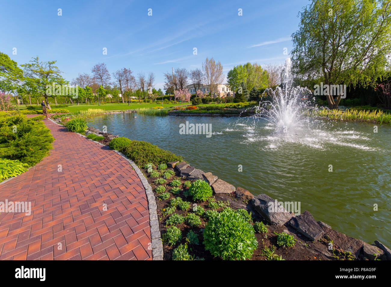 A path in the park passing next to a small pond with a fountain. With ...